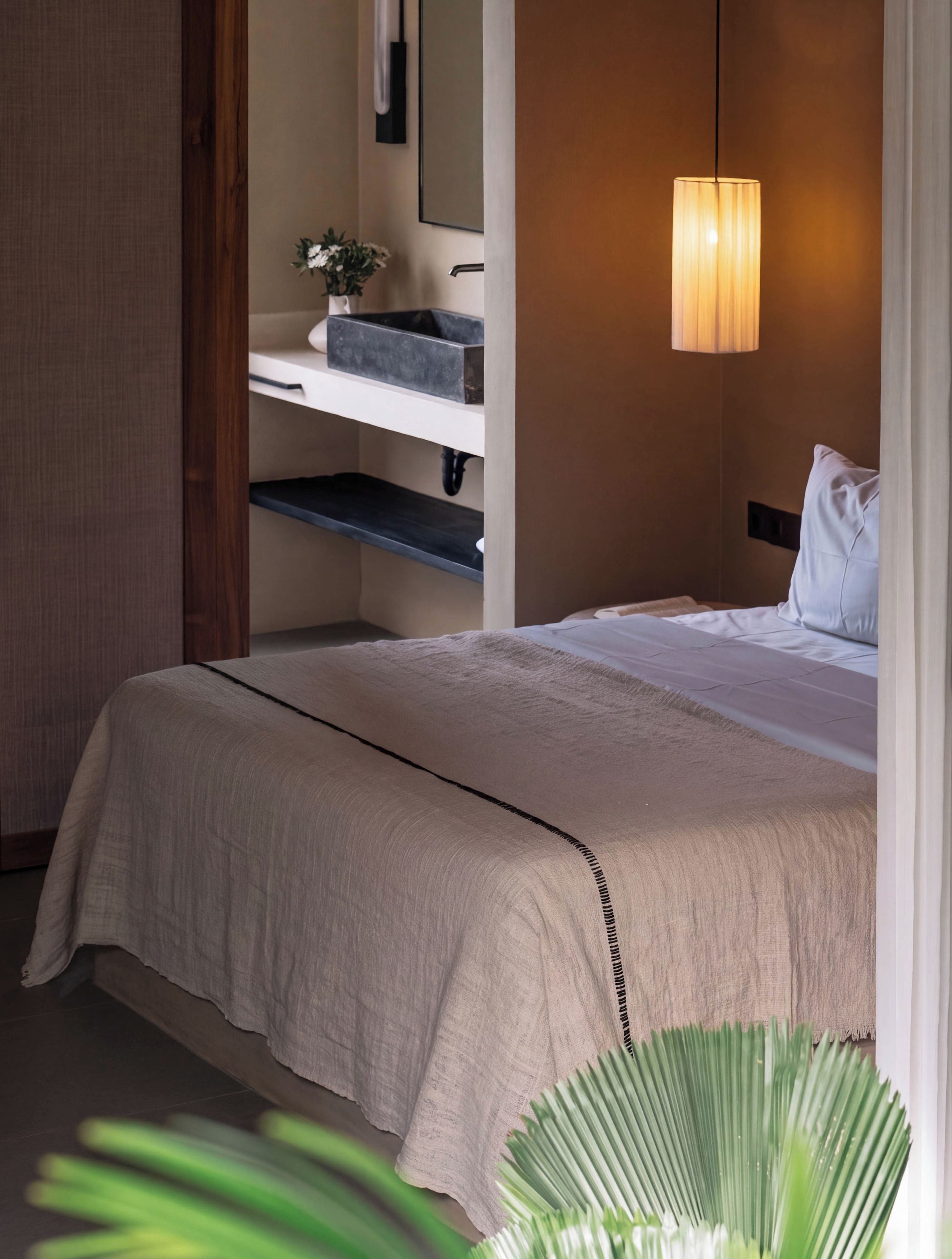 Quarto de hotel com cama, mesa de cabeceira, espelho, luminária pendente e uma planta.