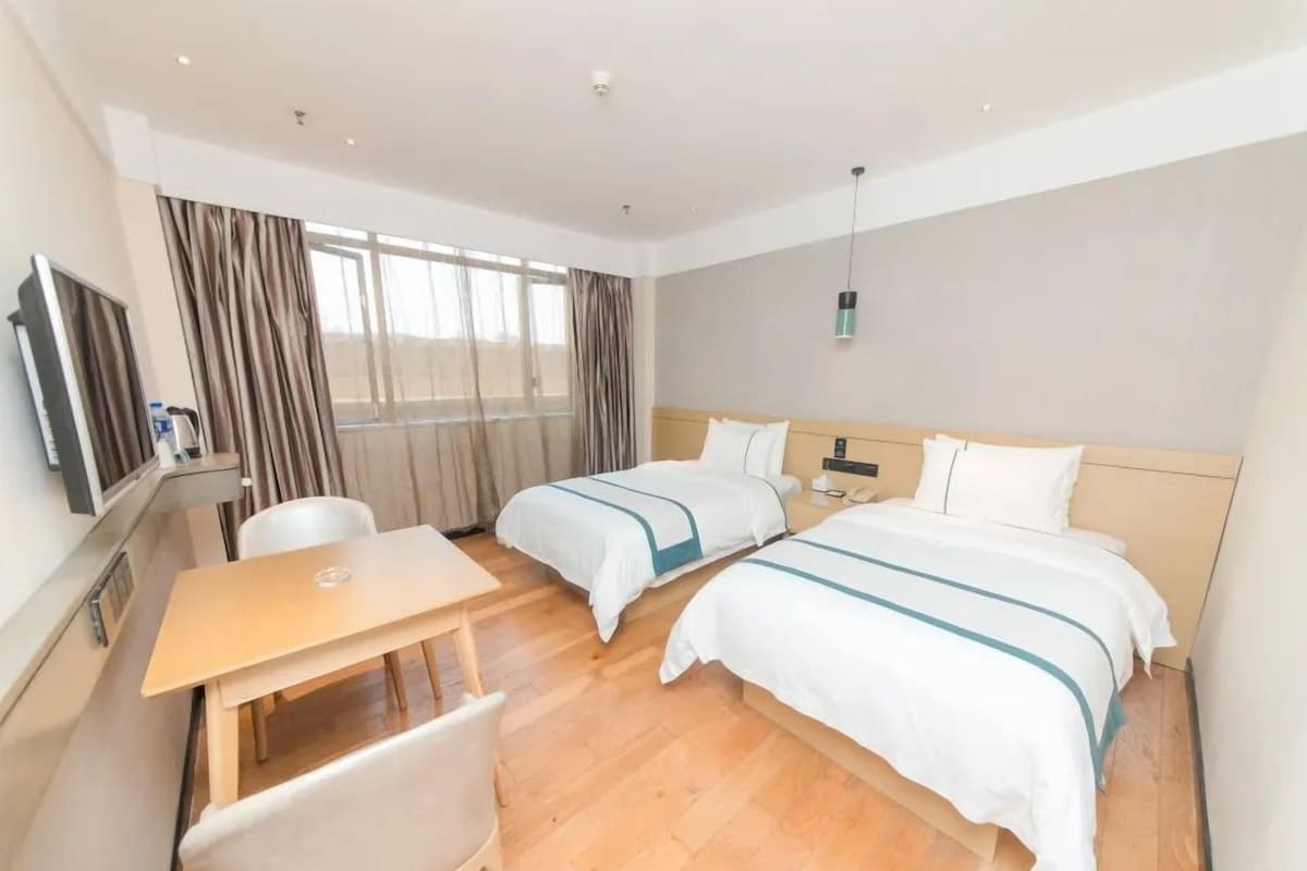 Quarto de hotel com duas camas, uma escrivaninha, uma cadeira, televisão e janela com cortinas.