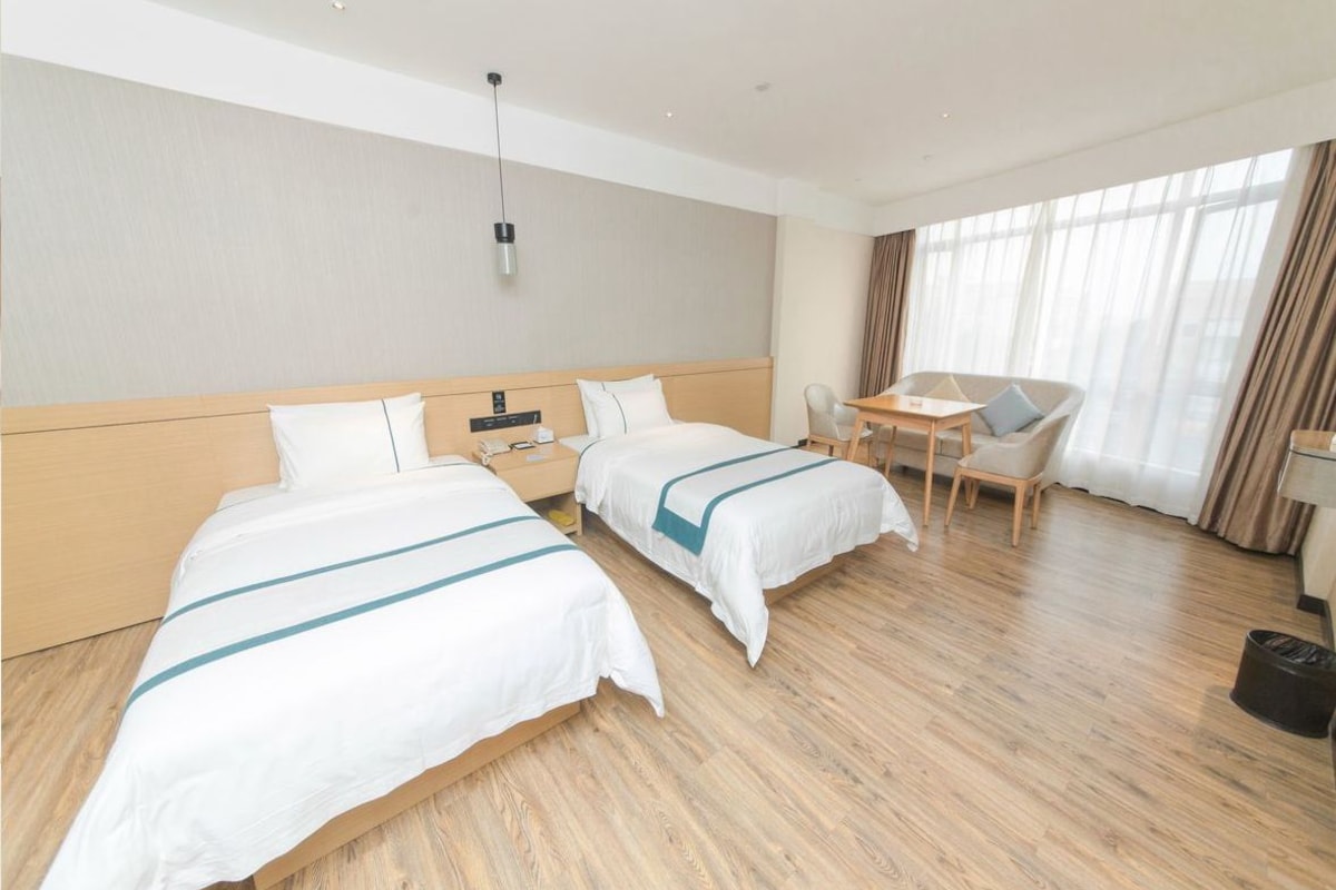 Quarto de hotel com duas camas, uma escrivaninha, uma cadeira e uma janela grande.
