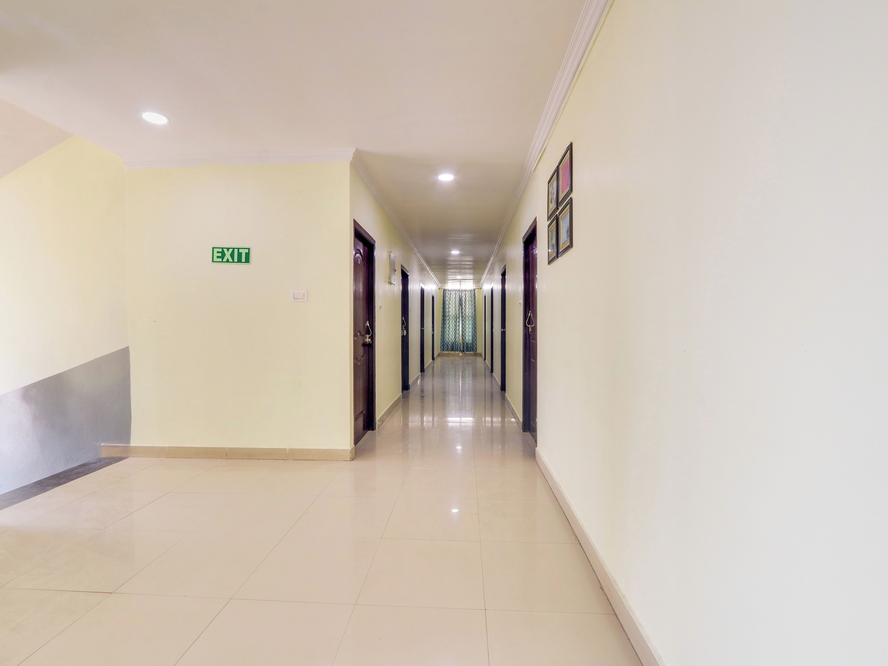Corredor longo e bem iluminado, com portas em ambos os lados, sinalização de saída e piso ladrilhado.
