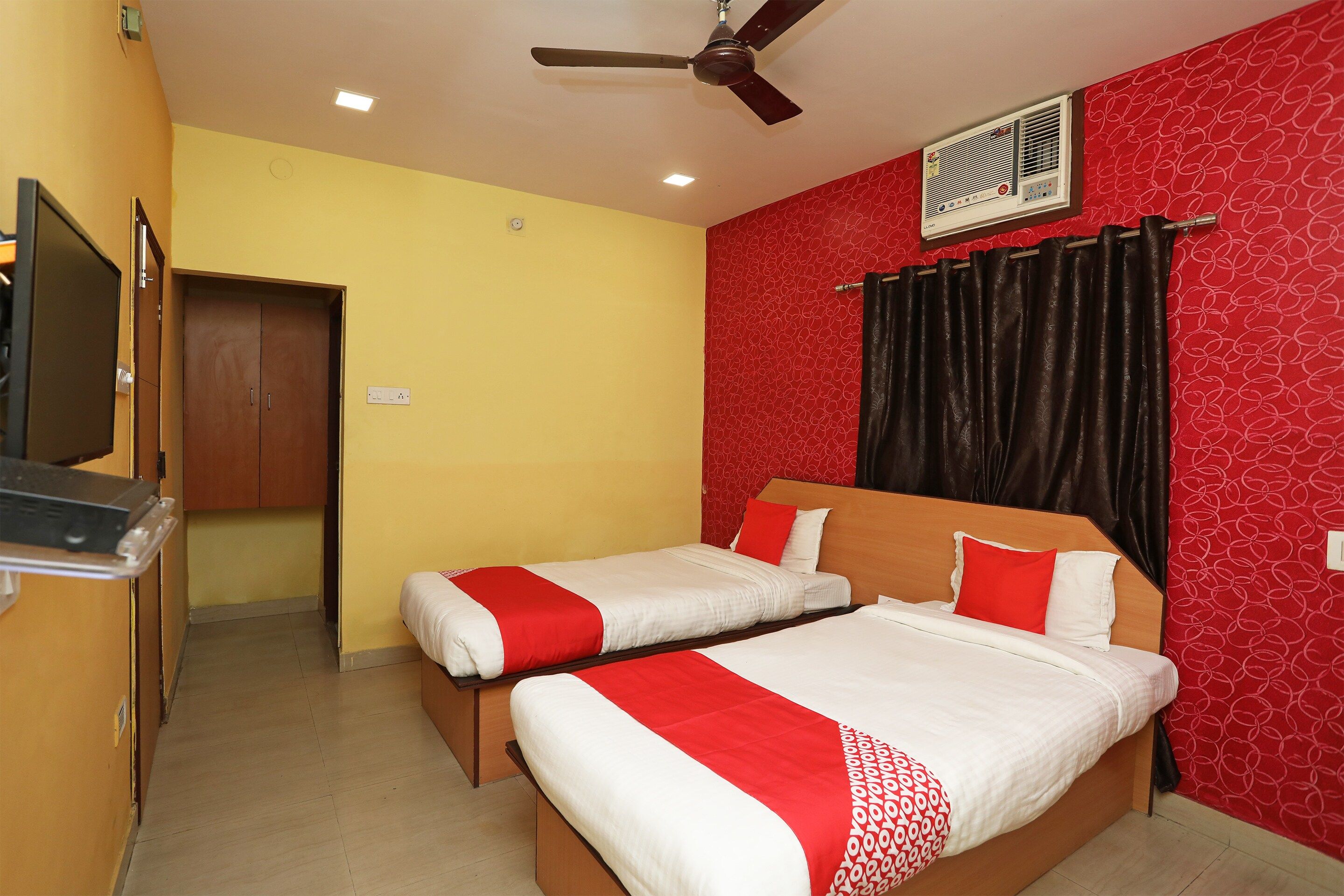 Quarto de hotel com duas camas, ventoinha no teto, ar condicionado, televisão e porta.