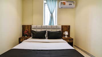 Quarto de hotel com cama, mesa de cabeceira com abajur, janela com cortinas e aparelho de ar condicionado na parede.