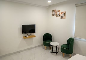 Quarto com televisão de tela plana na parede, duas poltronas estofadas em verde, uma mesa redonda pequena e uma prateleira de madeira abaixo da TV.