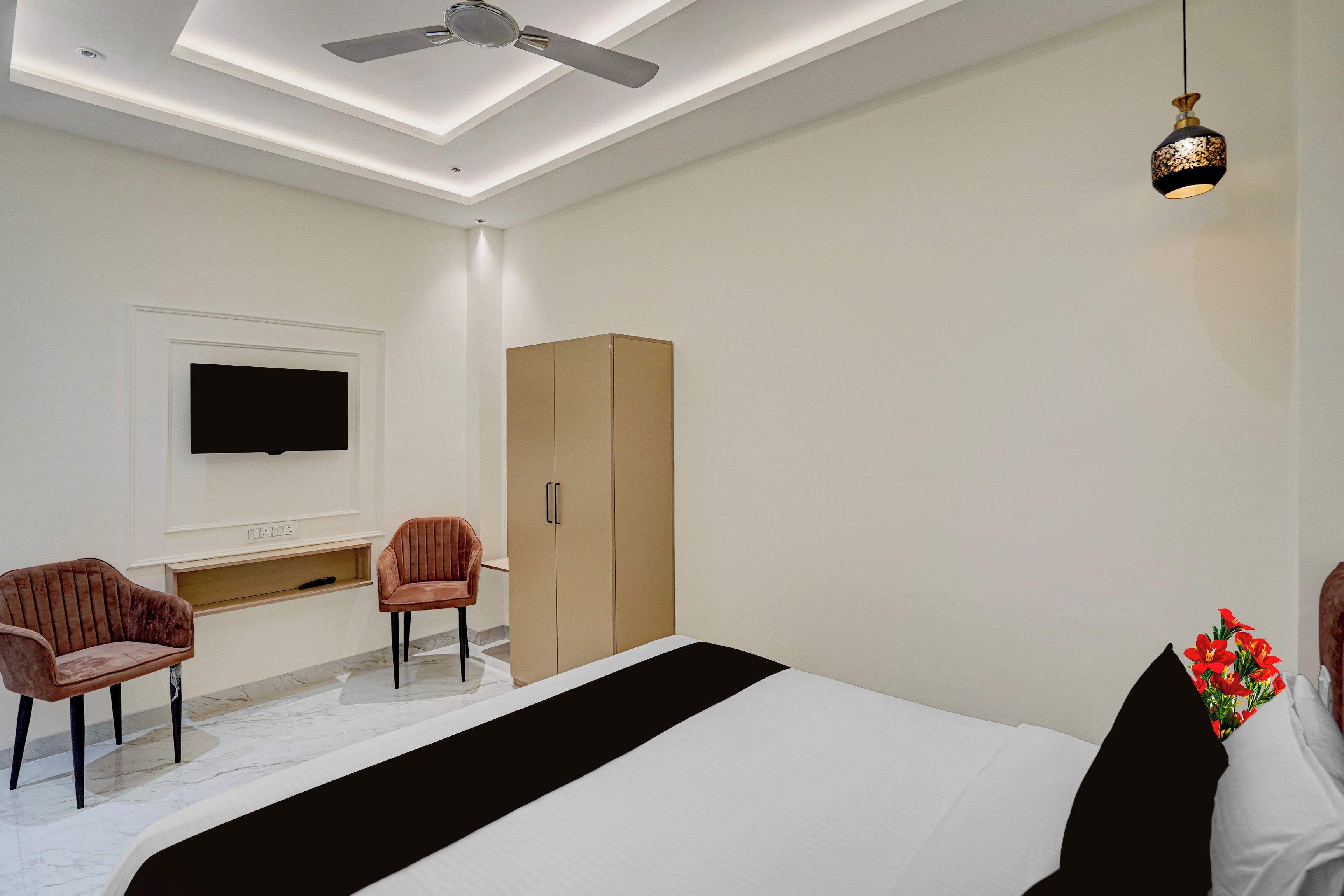 Quarto de hotel com cama, duas cadeiras, guarda-roupa, televisão e ventoinha no teto.