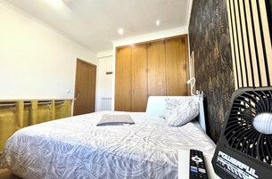 Quarto de hotel com cama, guarda-roupa de madeira, radiador e tomada.