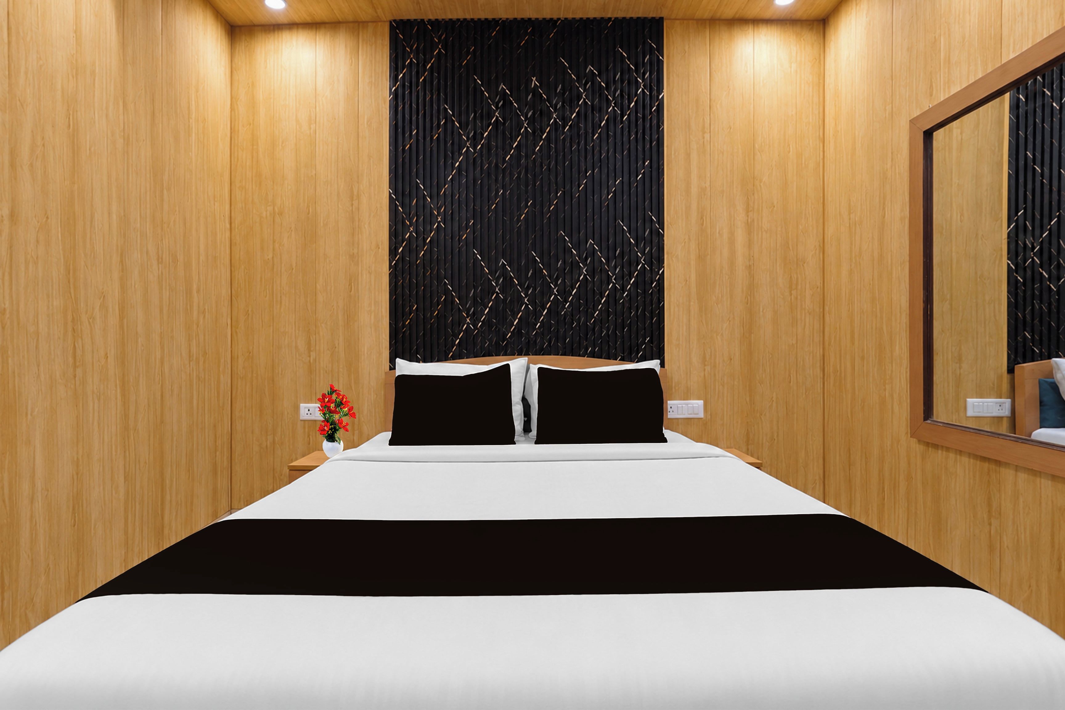 Quarto de hotel com uma cama grande, paredes de madeira, painel de parede preto e branco estampado, mesa de cabeceira com um vaso de flores e espelho na parede.