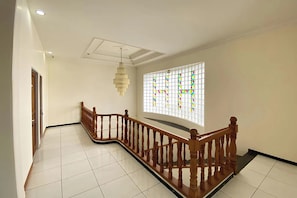 Uma escadaria com balaústres de madeira e corrimão, que leva a um patamar com um ventilador de teto e uma janela com padrão quadriculado.