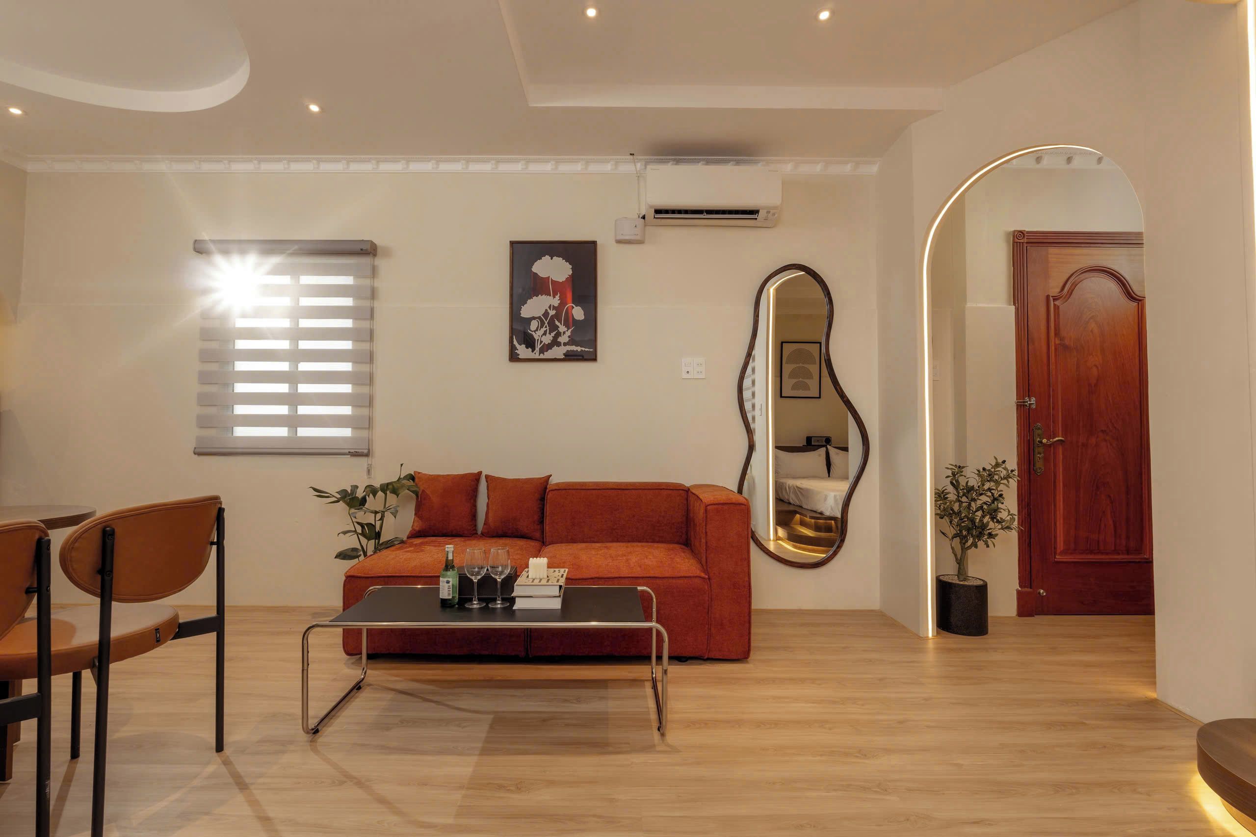 Sala de estar com sofá, mesa de centro e cadeiras. Há uma janela com persianas, um ar-condicionado fixado na parede e uma porta com espelho.
