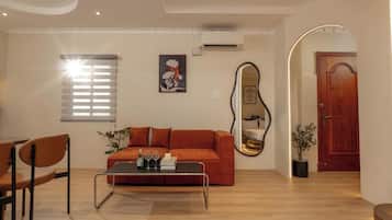 Sala de estar com sofá, mesa de centro e cadeiras. Há uma janela com persianas, um ar-condicionado fixado na parede e uma porta com espelho.