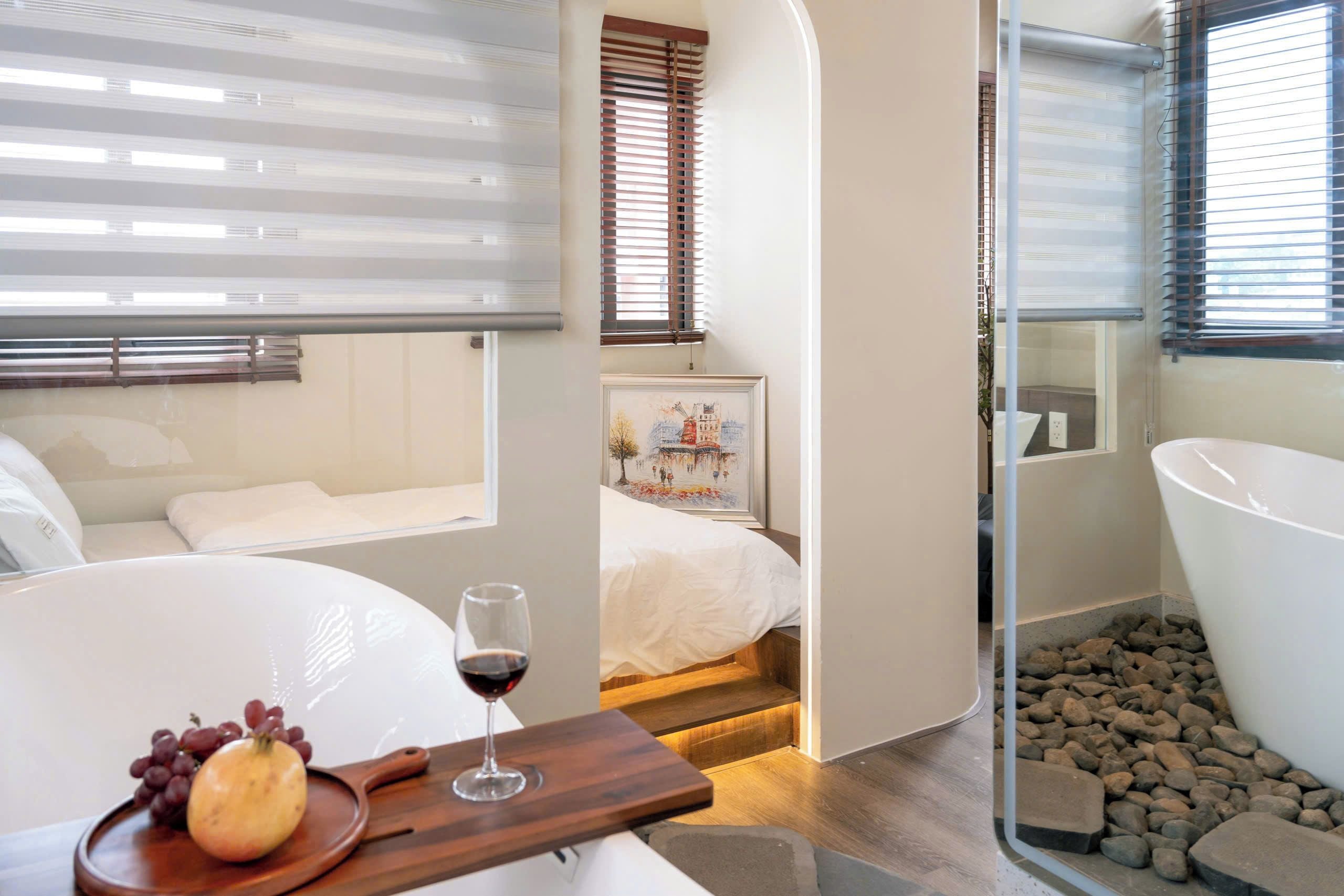 Quarto de hotel moderno com uma cama grande, banheiro com paredes de vidro e área de refeições com uma bandeja de frutas e taça de vinho.