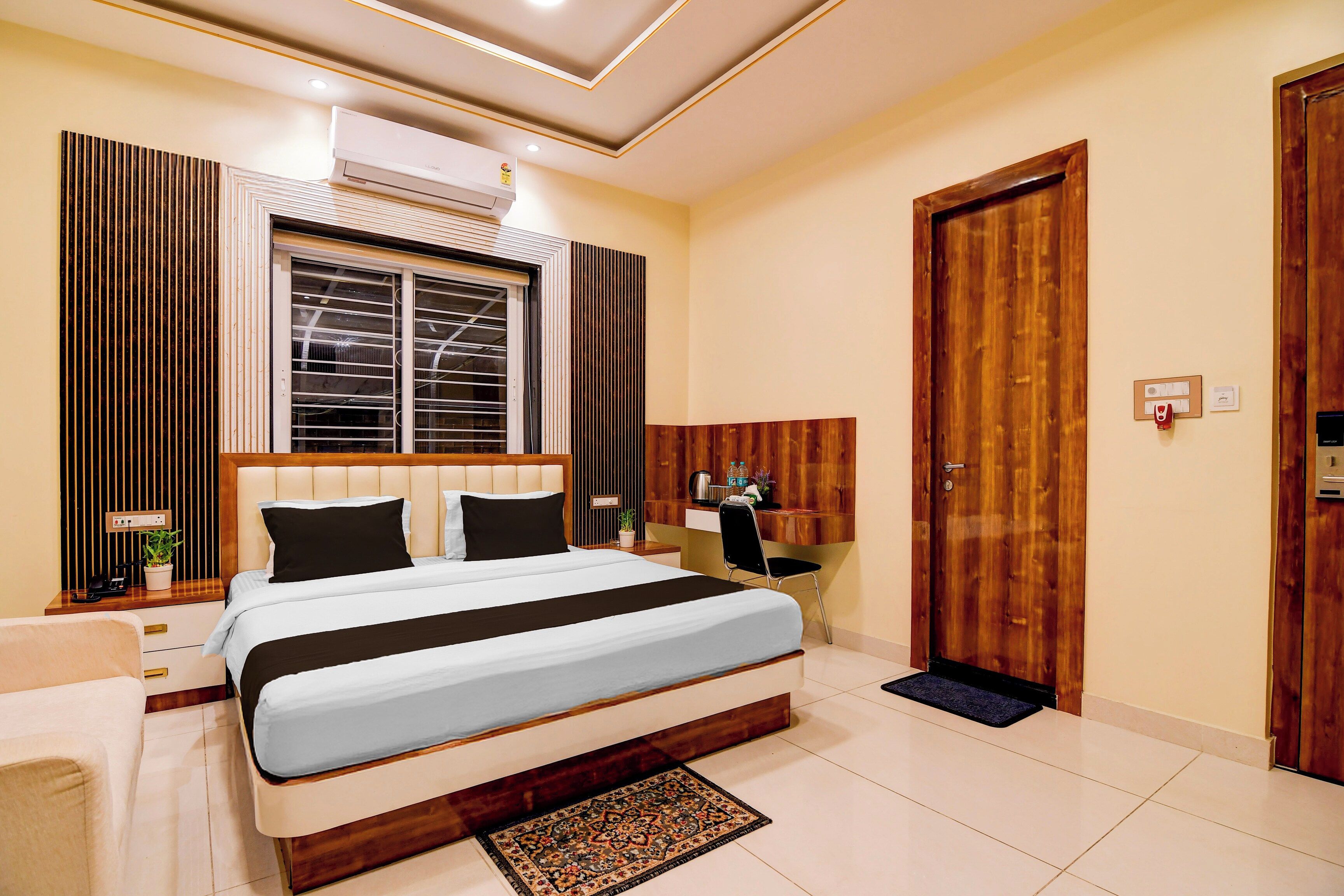 Quarto de hotel com cama, mesa de cabeceira, luminária, janela com persianas e porta de madeira.