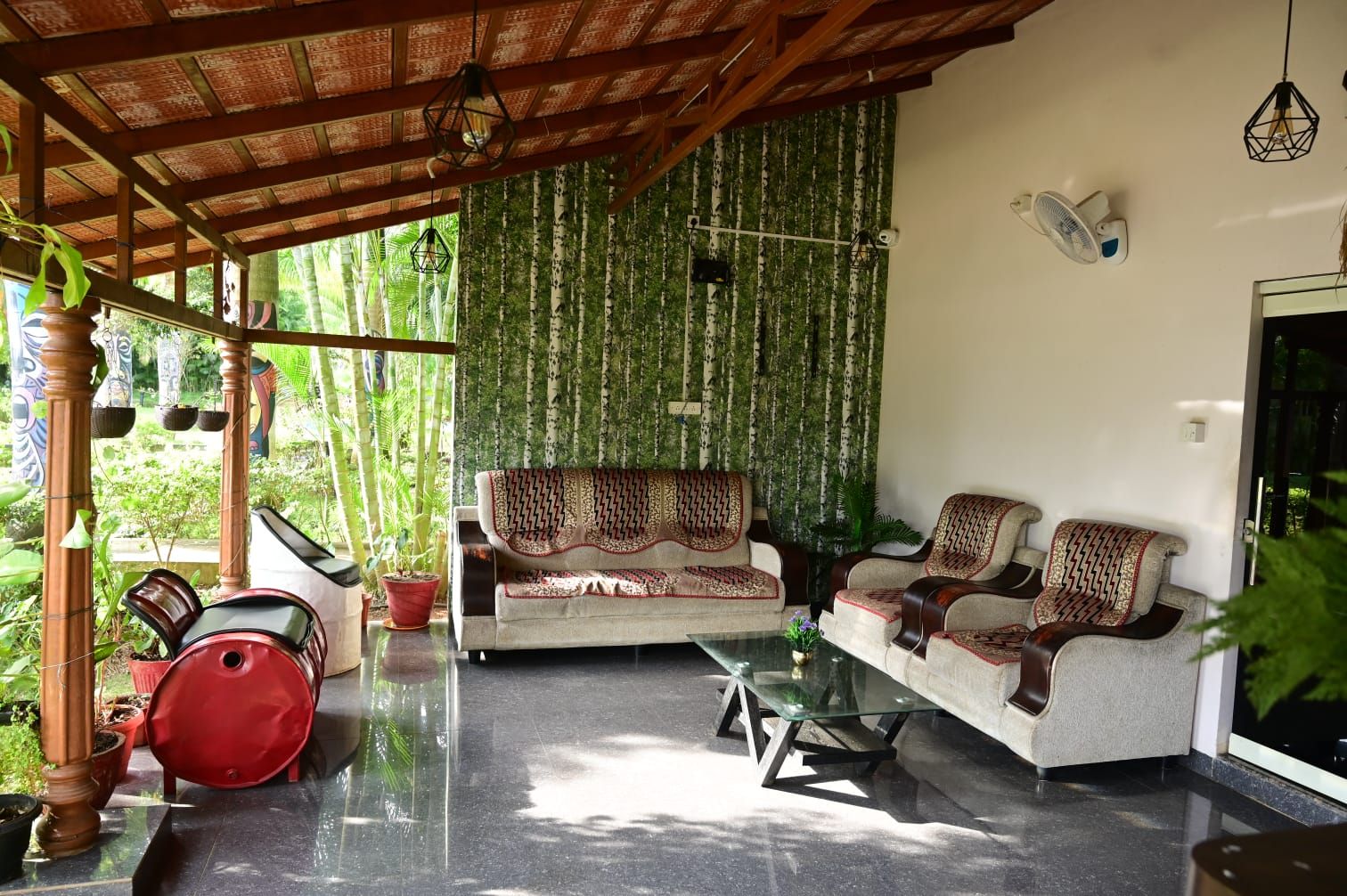 Uma área de estar coberta ao ar livre, com sofá, poltronas, mesa de centro de vidro e plantas em vasos.