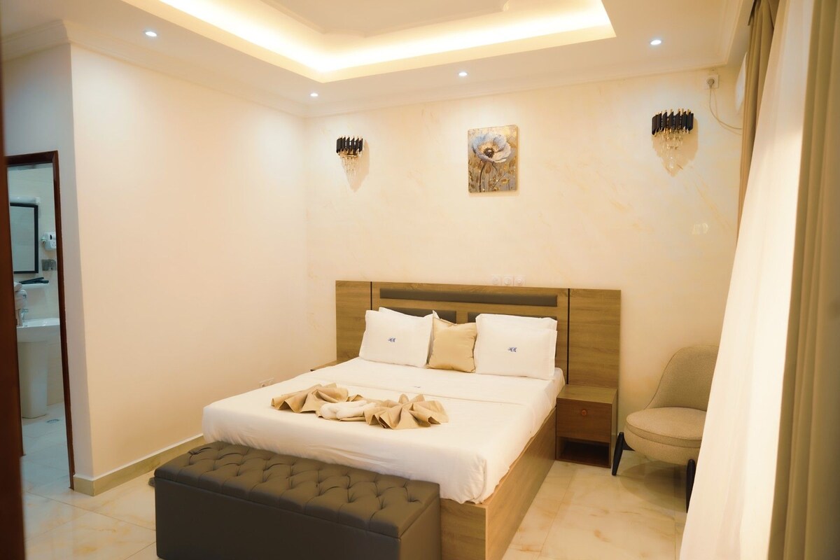 Quarto com cabeceira de madeira, cama com roupa de cama branca, mesa de cabeceira e uma cadeira.