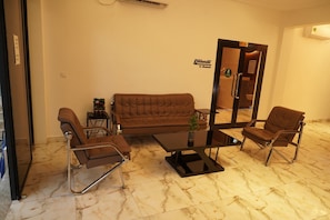 Quarto de hotel com sofá de couro marrom, duas poltronas, mesa de centro de vidro e piso de mármore.