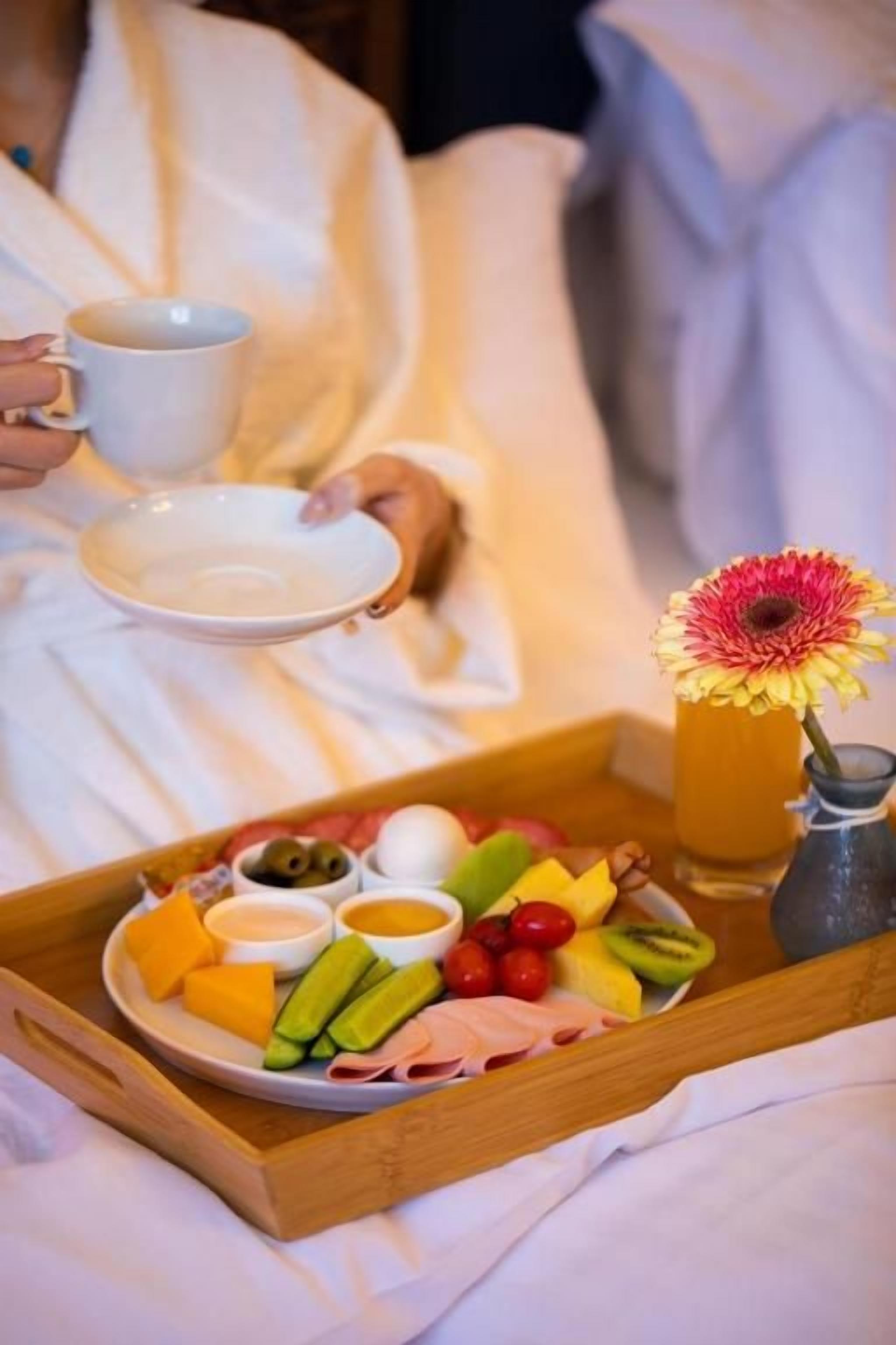 Uma bandeja com diversos alimentos, incluindo frutas, ovos e queijo, sobre uma cama, com uma pessoa segurando uma xícara e pires.