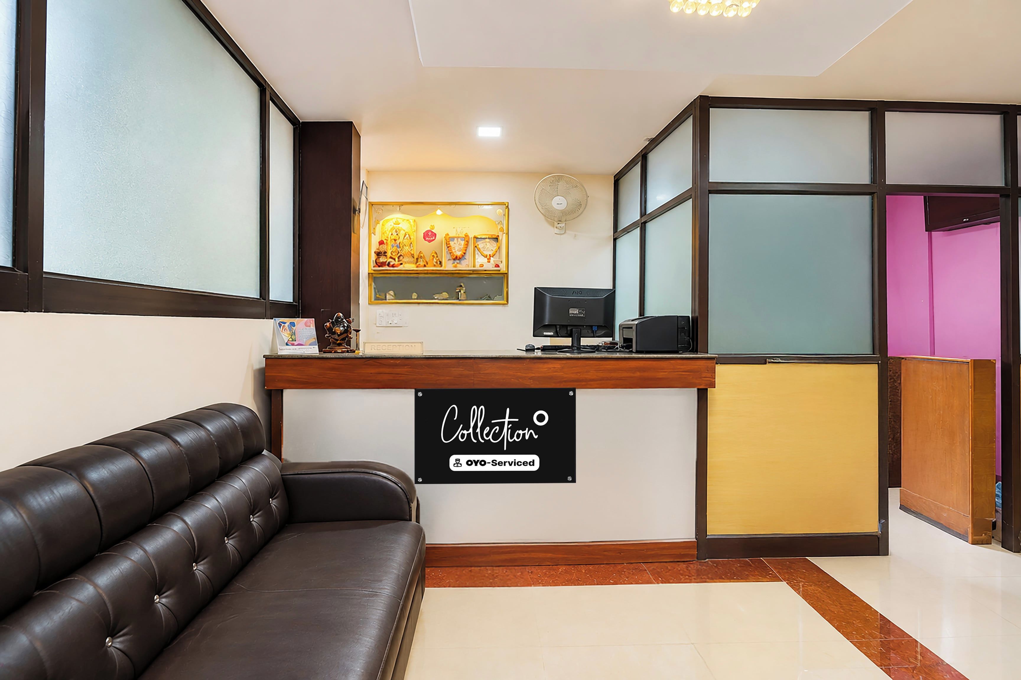 Uma área de recepção com uma placa preta indicando 'Collection OYO-Serviced', um balcão de atendimento, um computador, um ventilador, um sofá e uma parede com um objeto decorativo.
