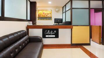 Uma área de recepção com uma placa preta indicando 'Collection OYO-Serviced', um balcão de atendimento, um computador, um ventilador, um sofá e uma parede com um objeto decorativo.