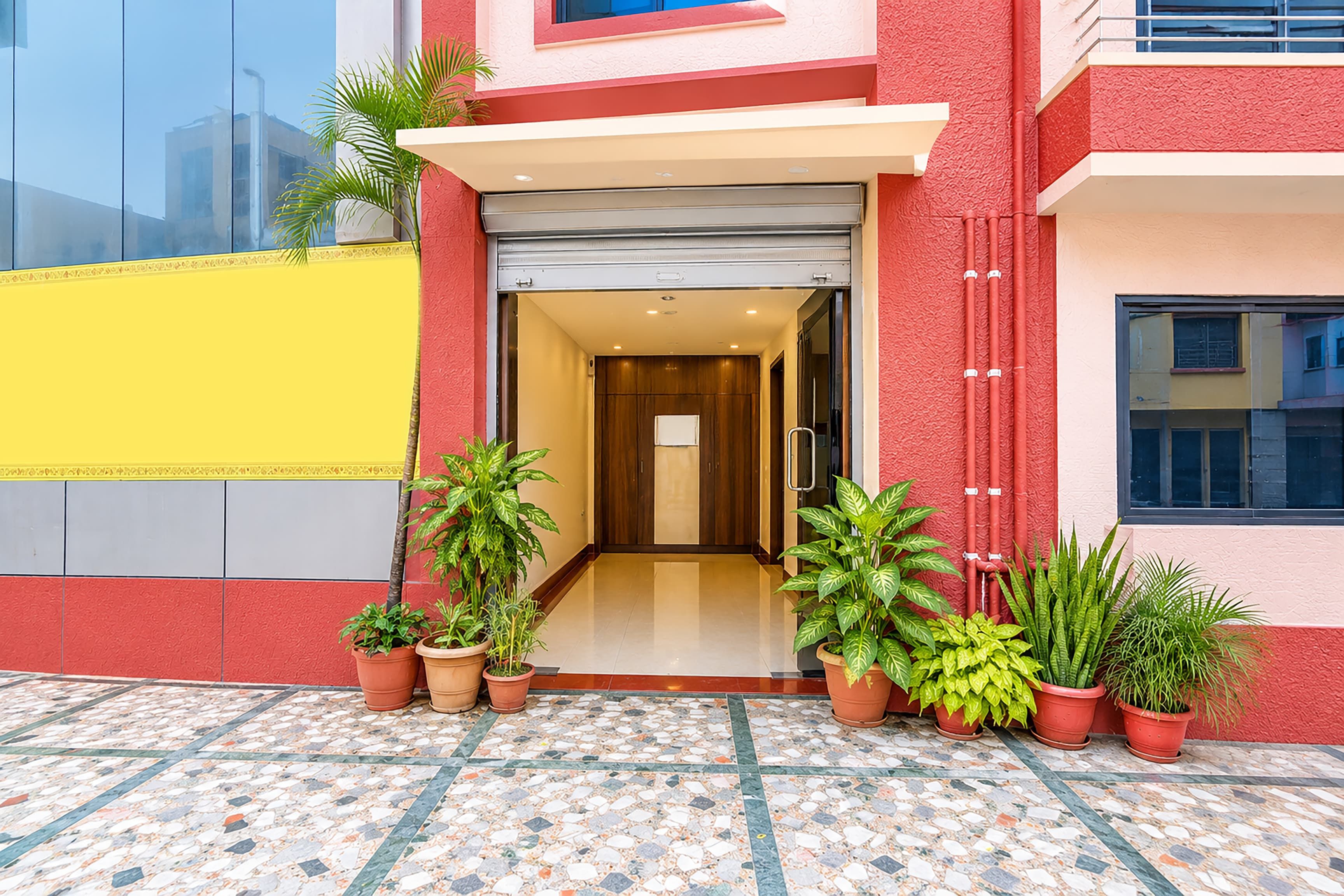 Entrada de hotel com piso de azulejos, plantas em vasos e fachada em vermelho e amarelo.