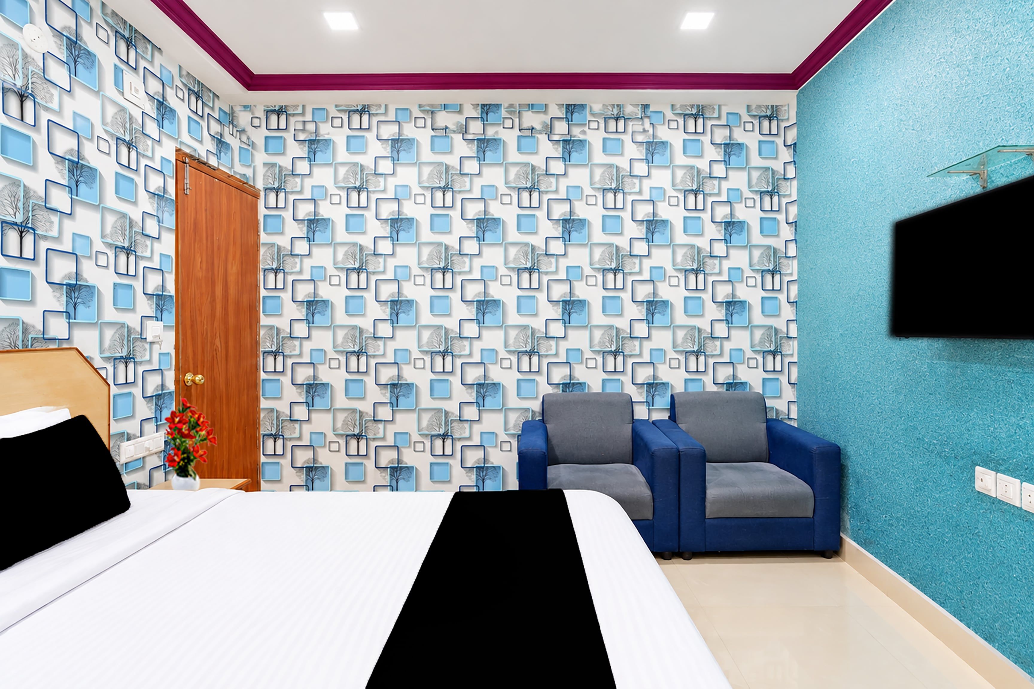 Quarto com papel de parede estampado, uma cama, duas poltronas, uma TV fixada na parede e uma porta de madeira.