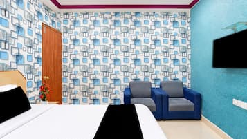 Quarto com papel de parede estampado, uma cama, duas poltronas, uma TV fixada na parede e uma porta de madeira.