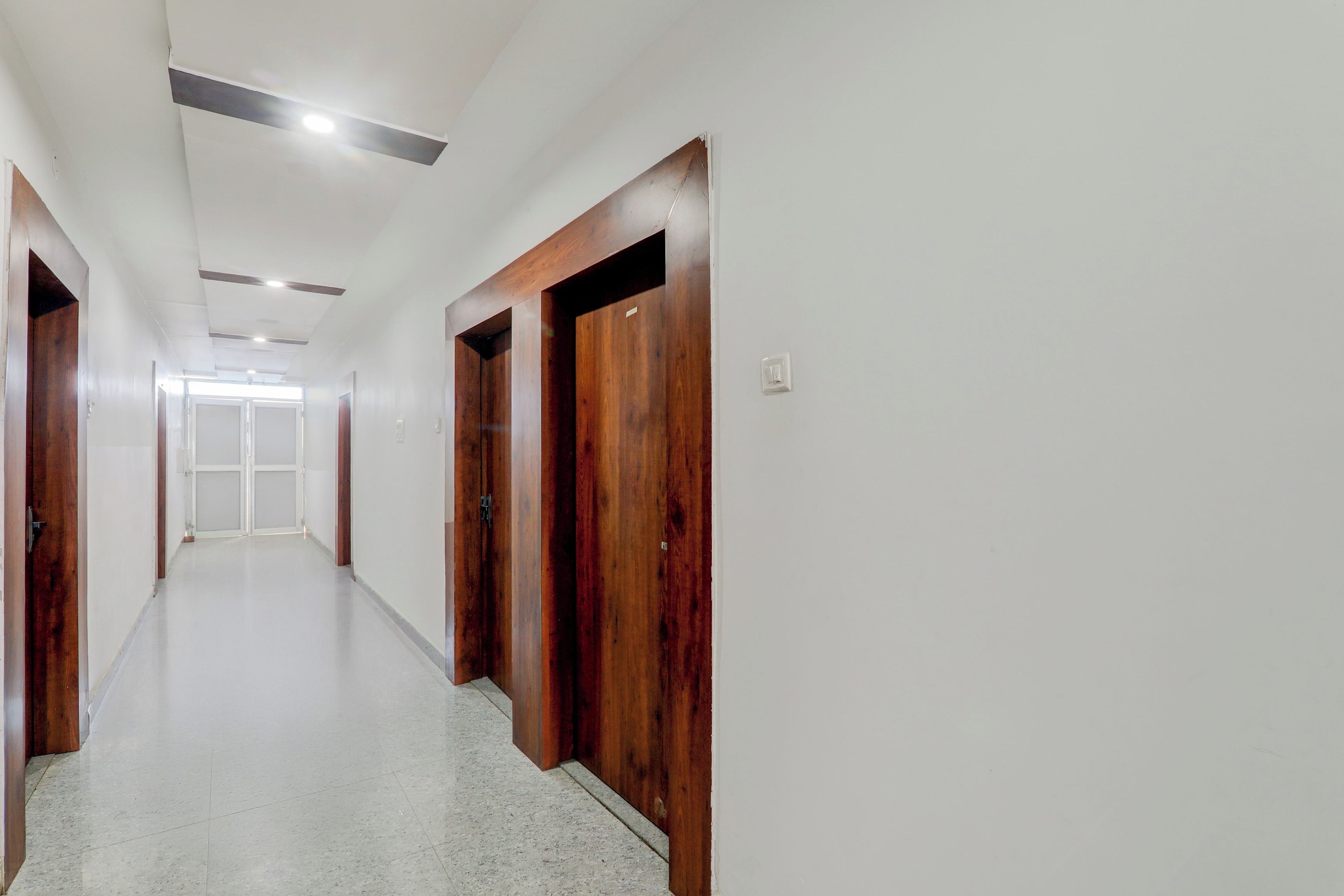 Um corredor com portas de madeira e paredes brancas, que leva a um quarto claro no final.