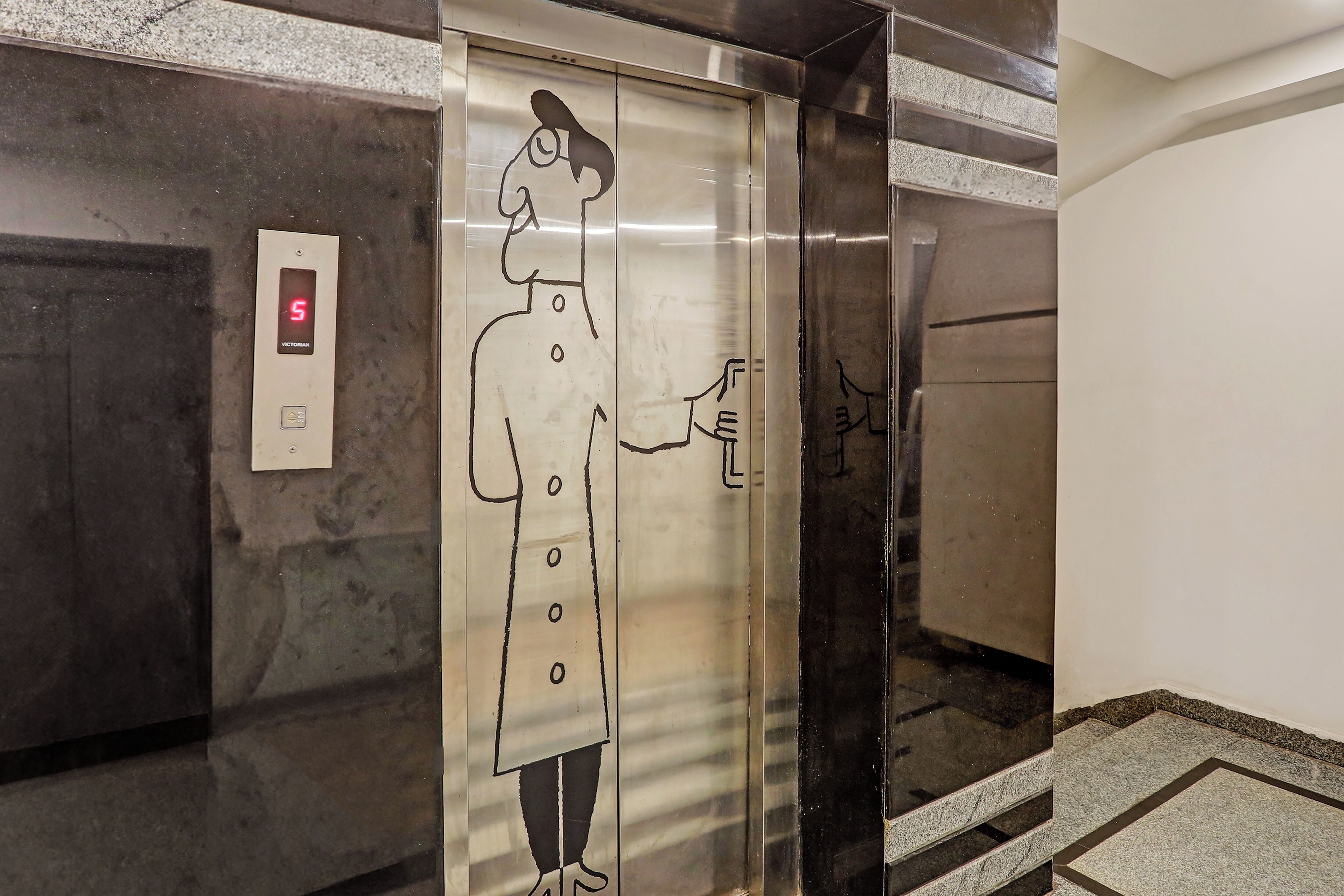 Interior de elevador com visor digital mostrando o número 5, ilustração de uma pessoa desenhada à mão e uma mão segurando um letreiro.