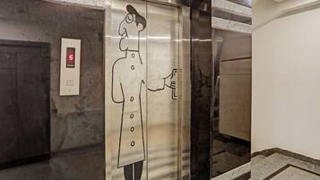 Interior de elevador com visor digital mostrando o número 5, ilustração de uma pessoa desenhada à mão e uma mão segurando um letreiro.