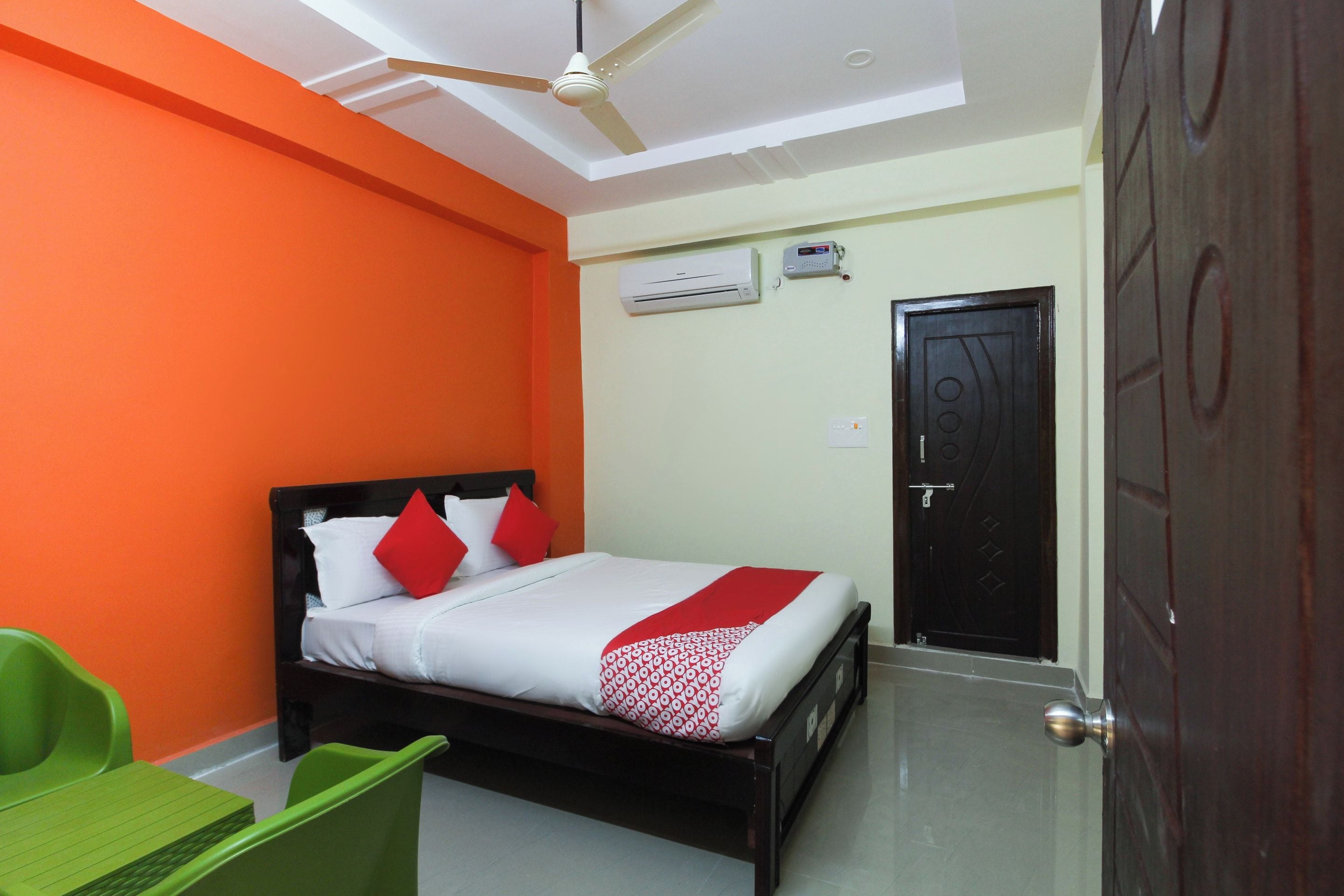 Quarto de hotel com cama, ventilador de teto, aparelho de ar condicionado, porta e uma cadeira verde.