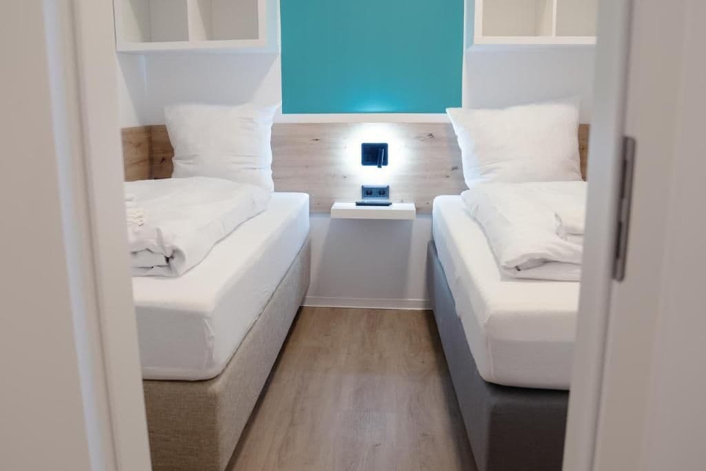 Quarto pequeno com duas camas, uma mesa e luminária na parede.