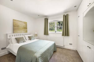 Quarto com cama, mesa de cabeceira, janela com cortinas, radiador e guarda-roupa.