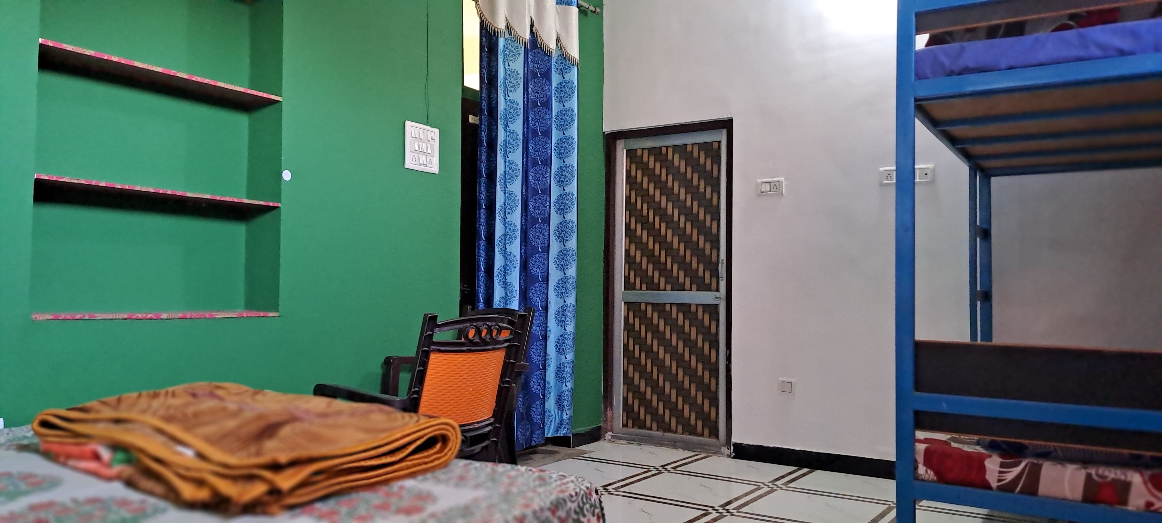 Quarto com uma cama de solteiro, uma cadeira, uma mesa com toalha estampada e uma porta com tapete padronizado.