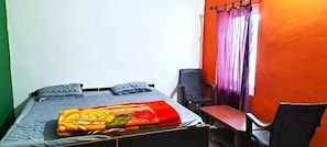 Uma cama de solteiro com colcha estampada, duas almofadas, uma cadeira e uma mesa pequena num quarto com paredes laranja e uma janela com cortinas.