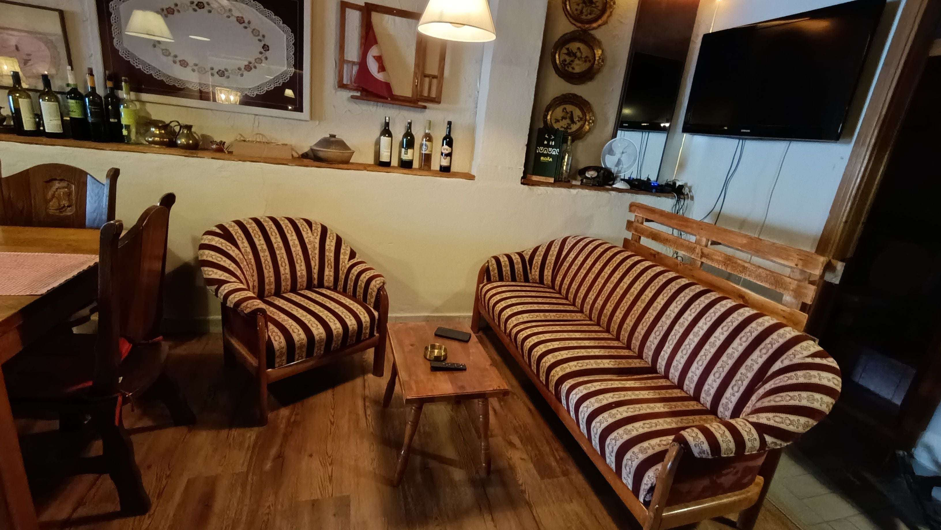Uma sala de estar com um sofá de madeira, uma poltrona listrada, uma mesa de centro de madeira e um espaço de jantar com mesa e cadeiras de madeira.