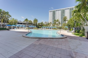 Piscina com espreguiçadeiras e guarda-sóis, cercada por palmeiras e um edifício alto ao fundo.