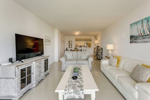 Uma sala de estar moderna com televisão de tela plana, um sofá branco, poltronas listradas e uma mesa de centro com toalha de mesa com tema de viagens. Uma cozinha aparece ao fundo.