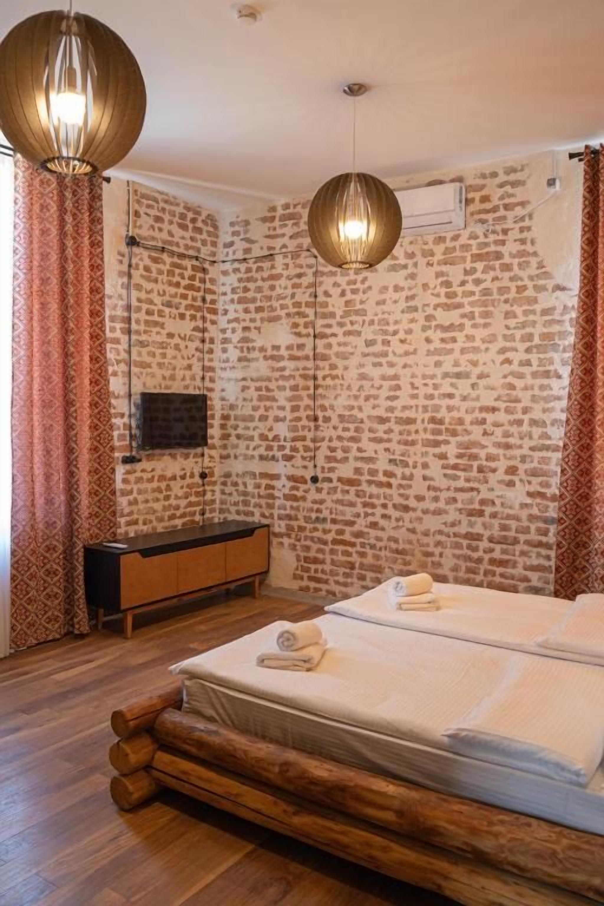 Quarto com parede de tijolos, cama com cabeceira de madeira, um banco de madeira e duas luminárias pendentes.