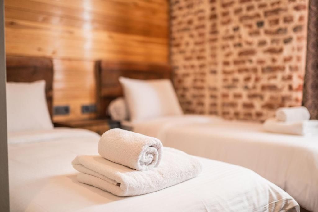 Quarto de hotel com duas camas, cada uma com uma toalha branca dobrada. O quarto tem paredes de madeira e uma parede de destaque com padrão de tijolos.