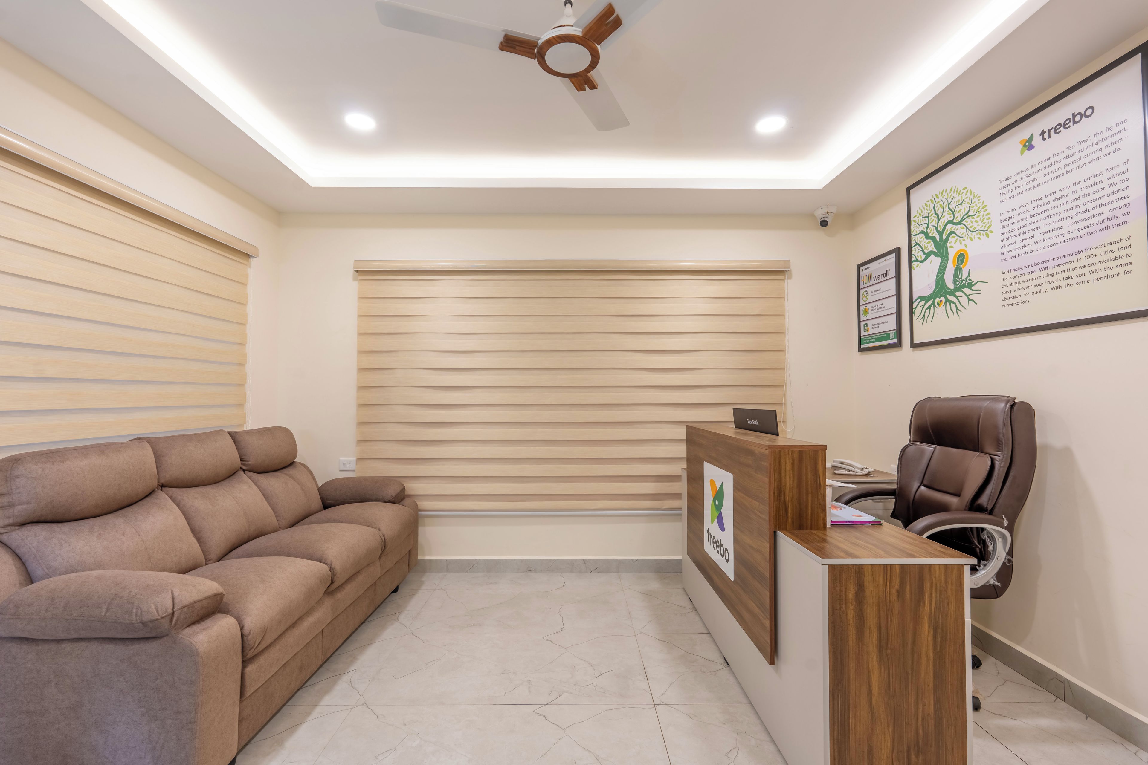 Uma área de recepção com sofá marrom, balcão de madeira, poltrona de couro marrom, ventilador de teto e placa fixada na parede.