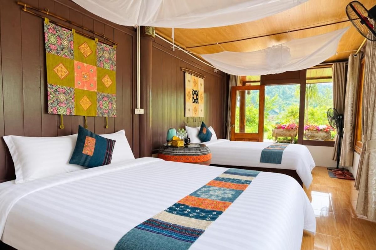 Quarto de hotel com duas camas, uma janela ampla com vista, mesa de cabeceira com abajur e colcha decorativa.