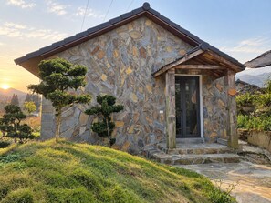 Uma casa de pedra com varanda de madeira e porta, cercada por vegetação e uma paisagem montanhosa.