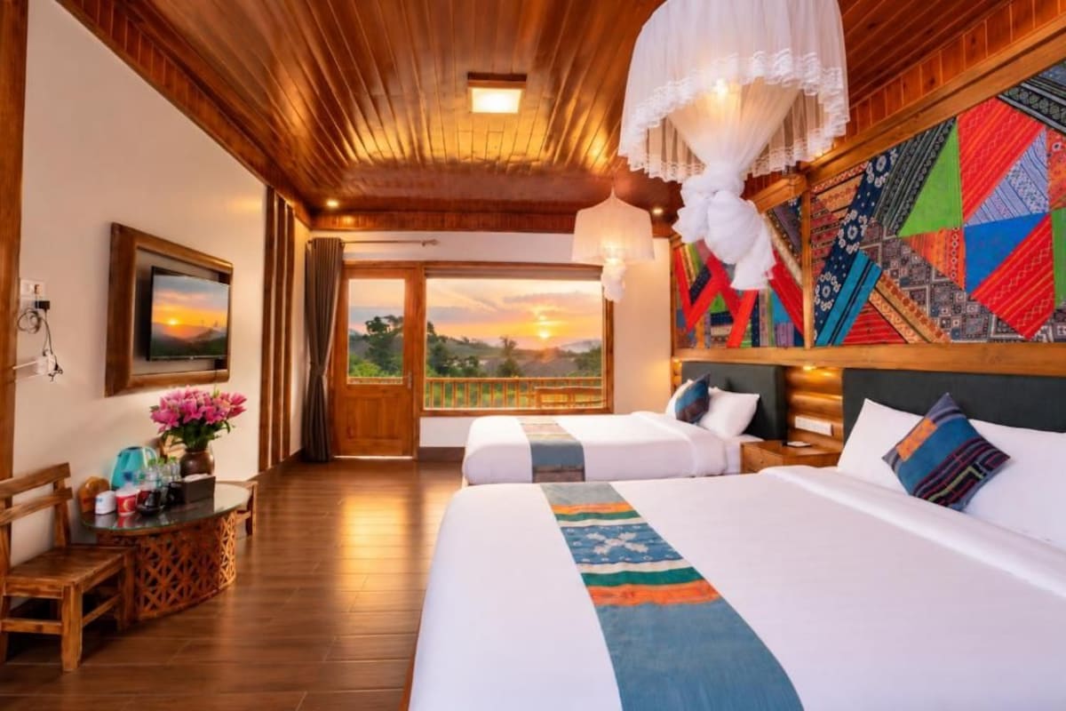 Quarto de hotel com cama grande, mesa de cabeceira, cadeira, janela com vista para o pôr do sol e parede com padrão geométrico colorido.