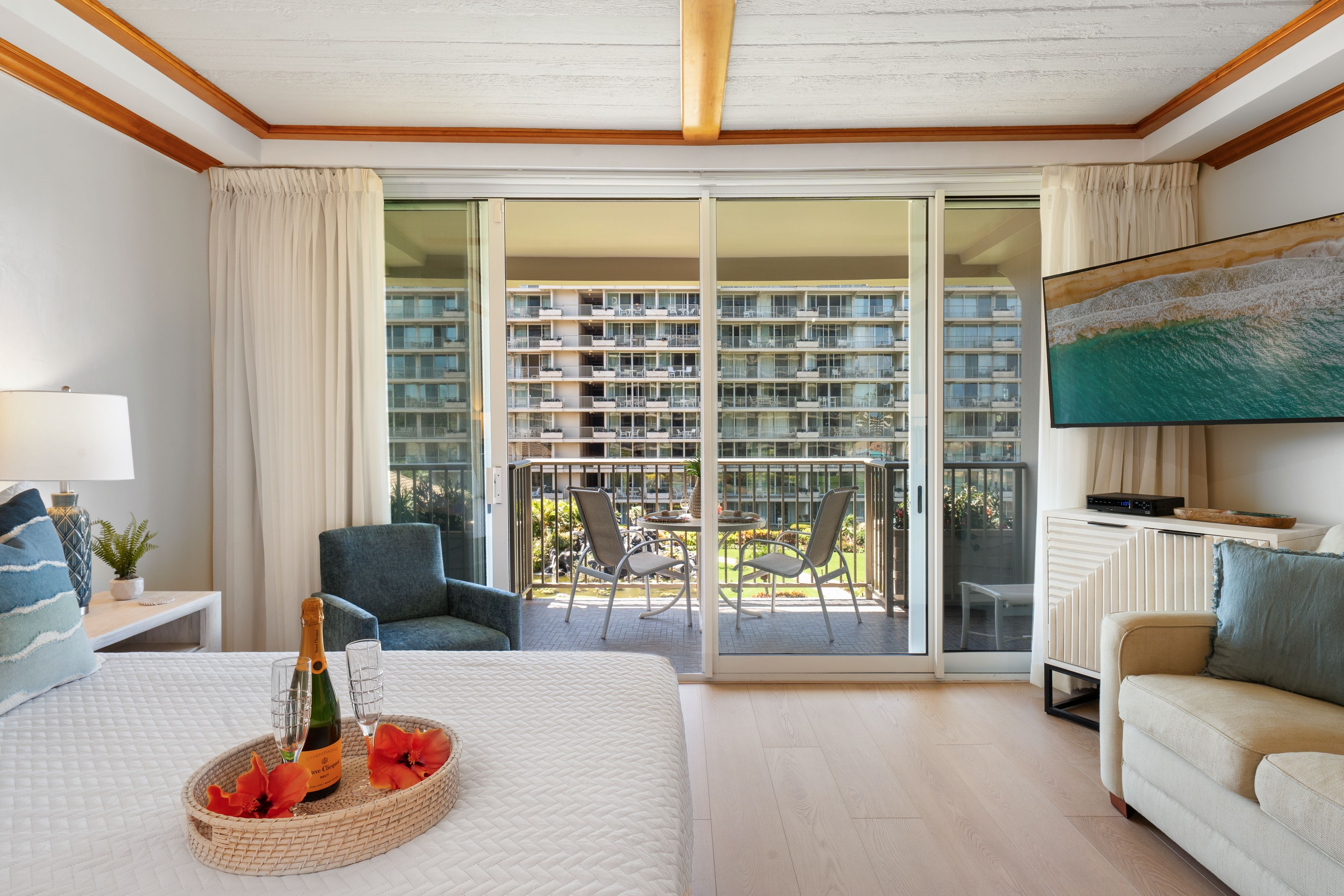 Quarto de hotel com cama, varanda e vista para outros edifícios, televisão na parede e mesa com garrafa e copos.
