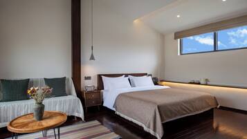 Quarto de hotel moderno com uma cama grande, um sofá, uma mesa pequena com um vaso de flores e uma janela com vista para o céu.