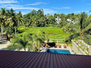 Área de piscina no terraço com espreguiçadeiras e um guarda-sol de palha, rodeada por vegetação exuberante e palmeiras.