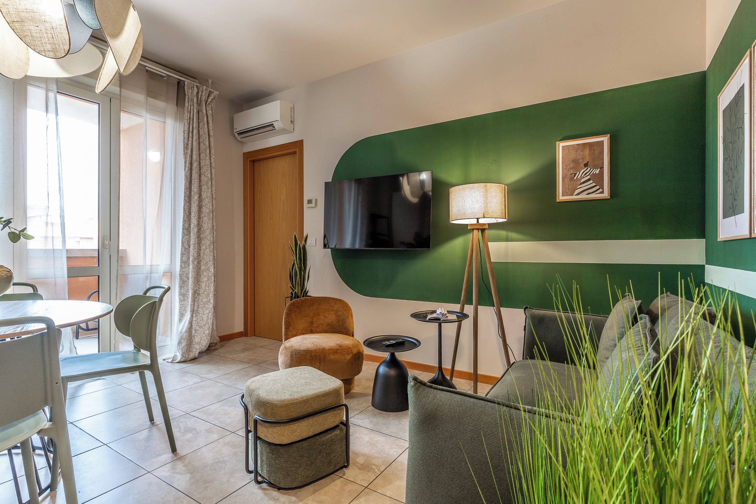 Uma sala de estar moderna com uma parede verde decorativa, uma TV de tela plana, um sofá, uma mesa de centro e uma área de jantar com cadeiras e uma mesa.