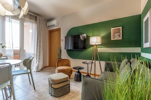 Uma sala de estar moderna com uma parede verde decorativa, uma TV de tela plana, um sofá, uma mesa de centro e uma área de jantar com cadeiras e uma mesa.