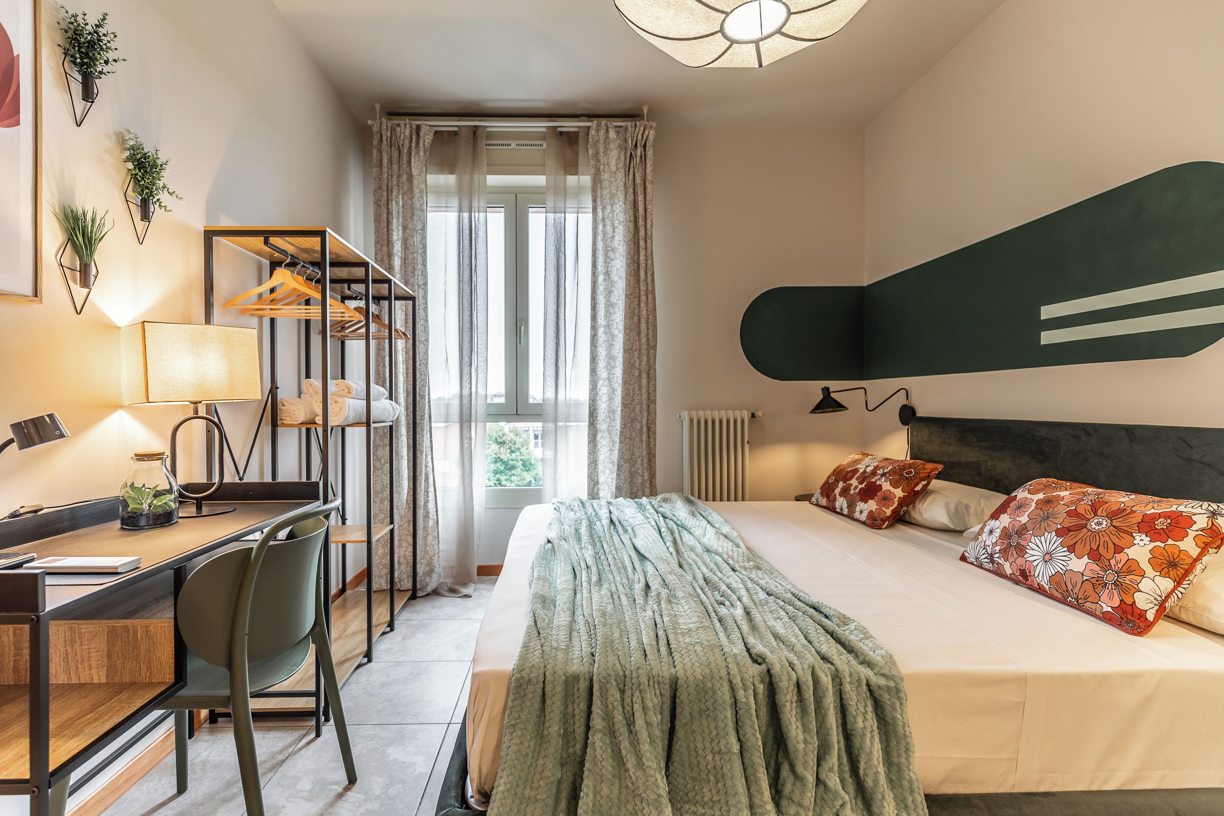 Quarto de hotel com cama, escrivaninha, cadeira, luminária e radiador.