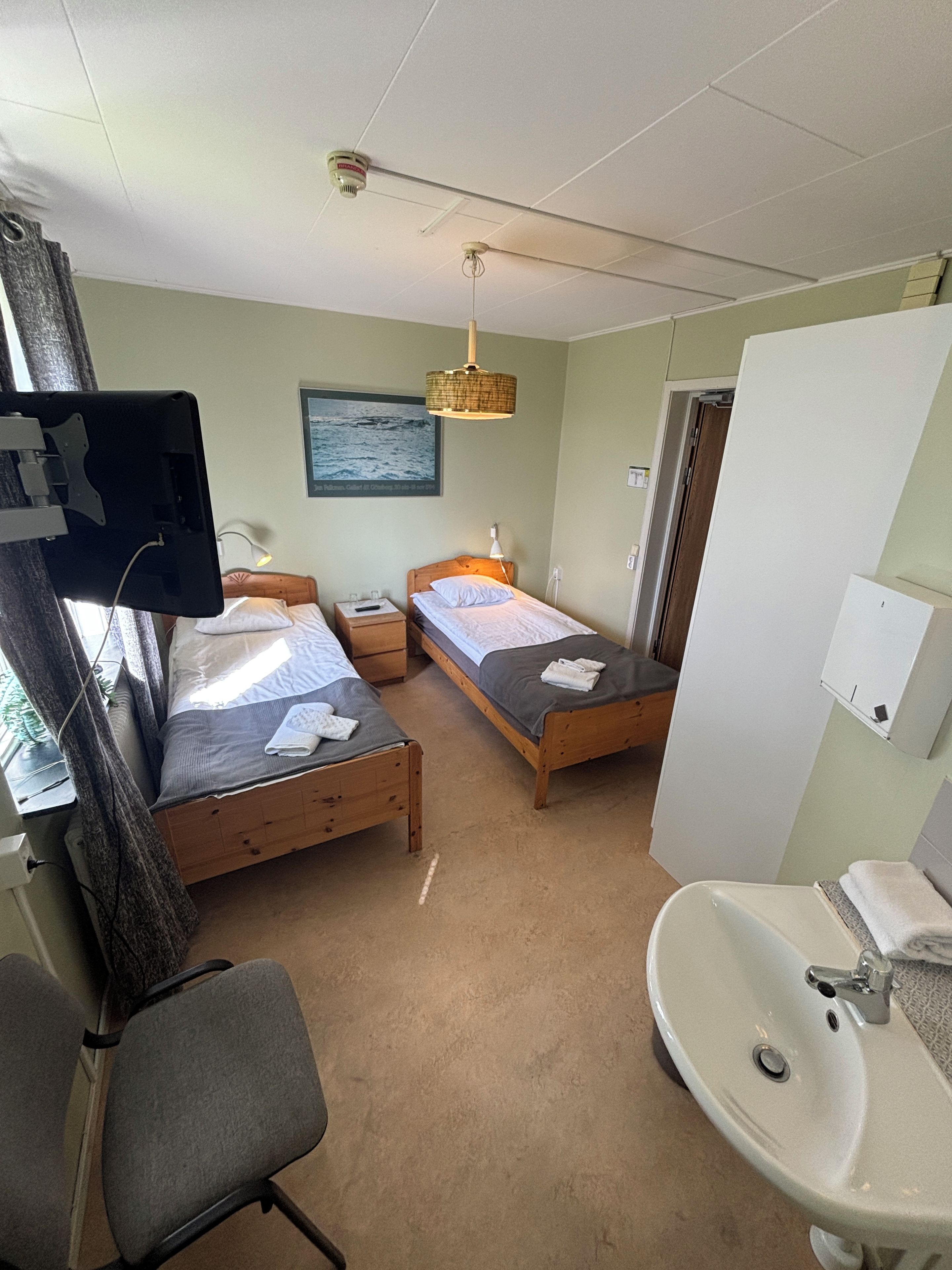 Quarto de hotel com duas camas de madeira, uma escrivaninha com cadeira, uma janela grande com cortinas, uma televisão na parede, um ventilador de teto e banheiro com pia e espelho.