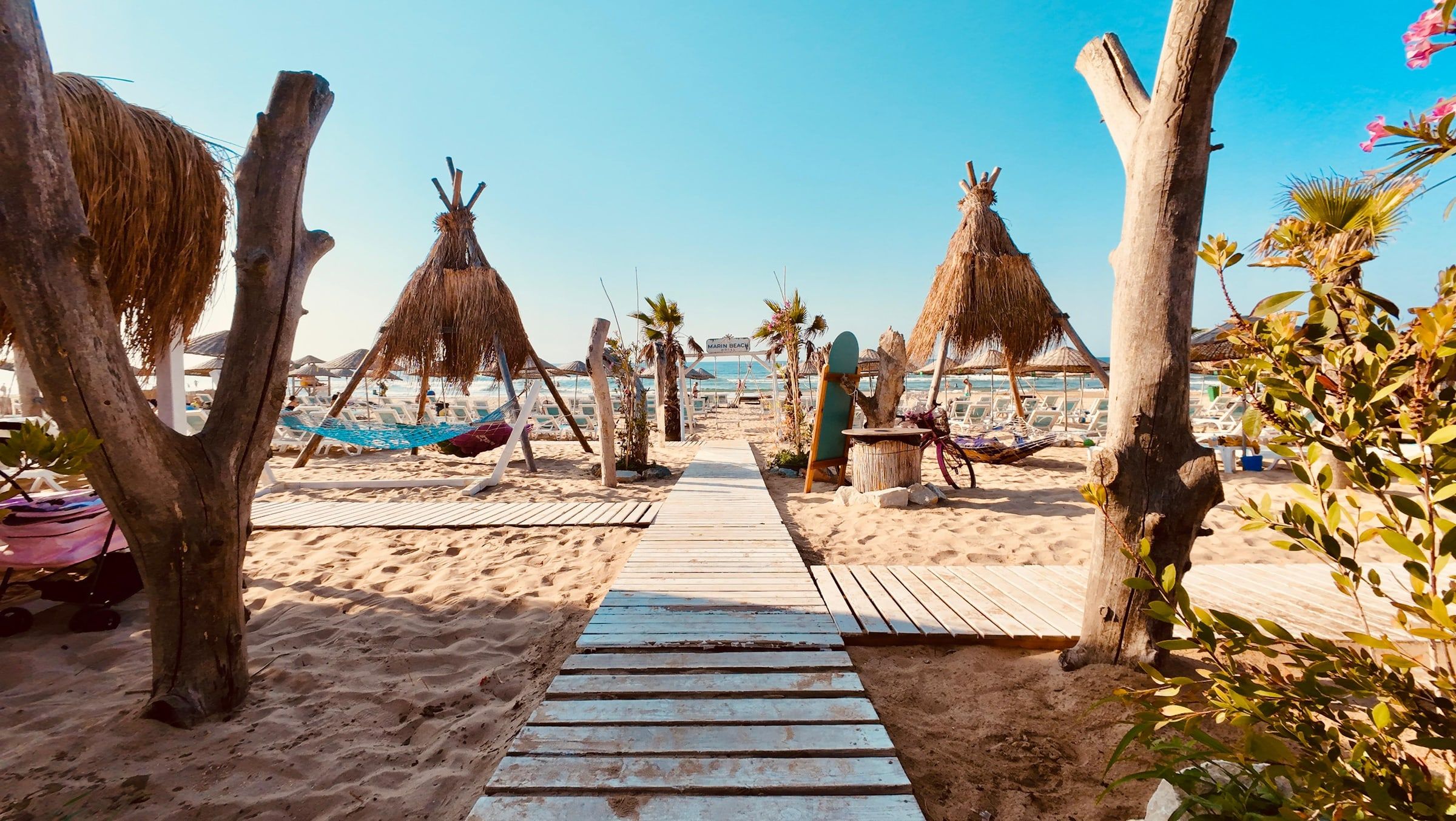 Uma trilha na praia que leva a uma área arenosa com guarda-sóis de palha e espreguiçadeiras.