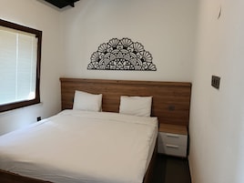 Quarto com cabeceira de madeira, cama com roupa de cama branca, mesa de cabeceira e um objeto decorativo na parede.